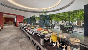 Buffet com diversos alimentos e um espaço para café num hall moderno de hotel, com amplas janelas e plantas internas.