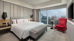 Quarto de hotel moderno com uma cama grande, uma poltrona vermelha, vista para a cidade e televisão de tela plana.