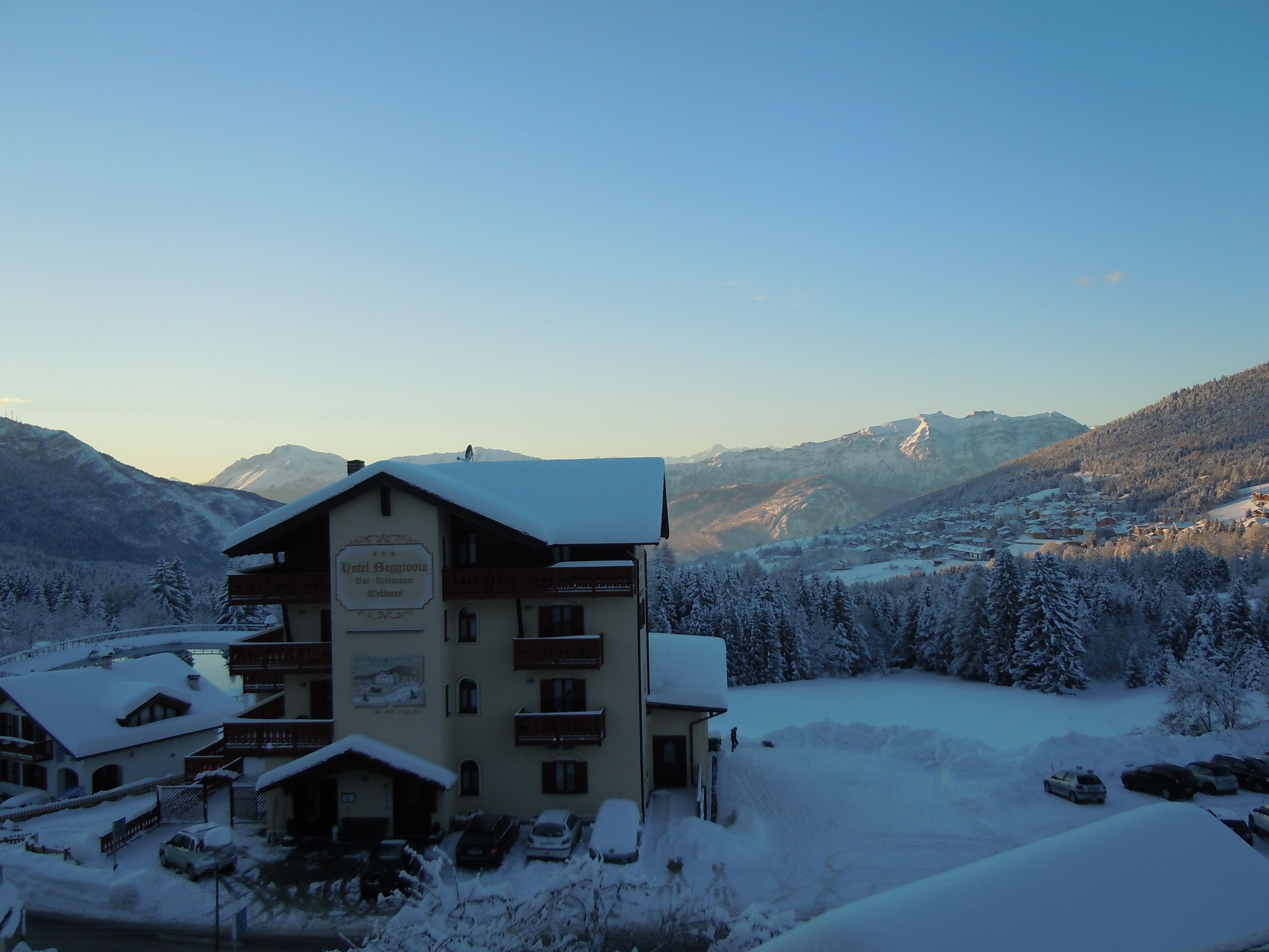 Um hotel com vários andares, com uma placa indicando ser um bar, restaurante e centro de bem-estar, rodeado por árvores cobertas de neve e montanhas ao fundo.