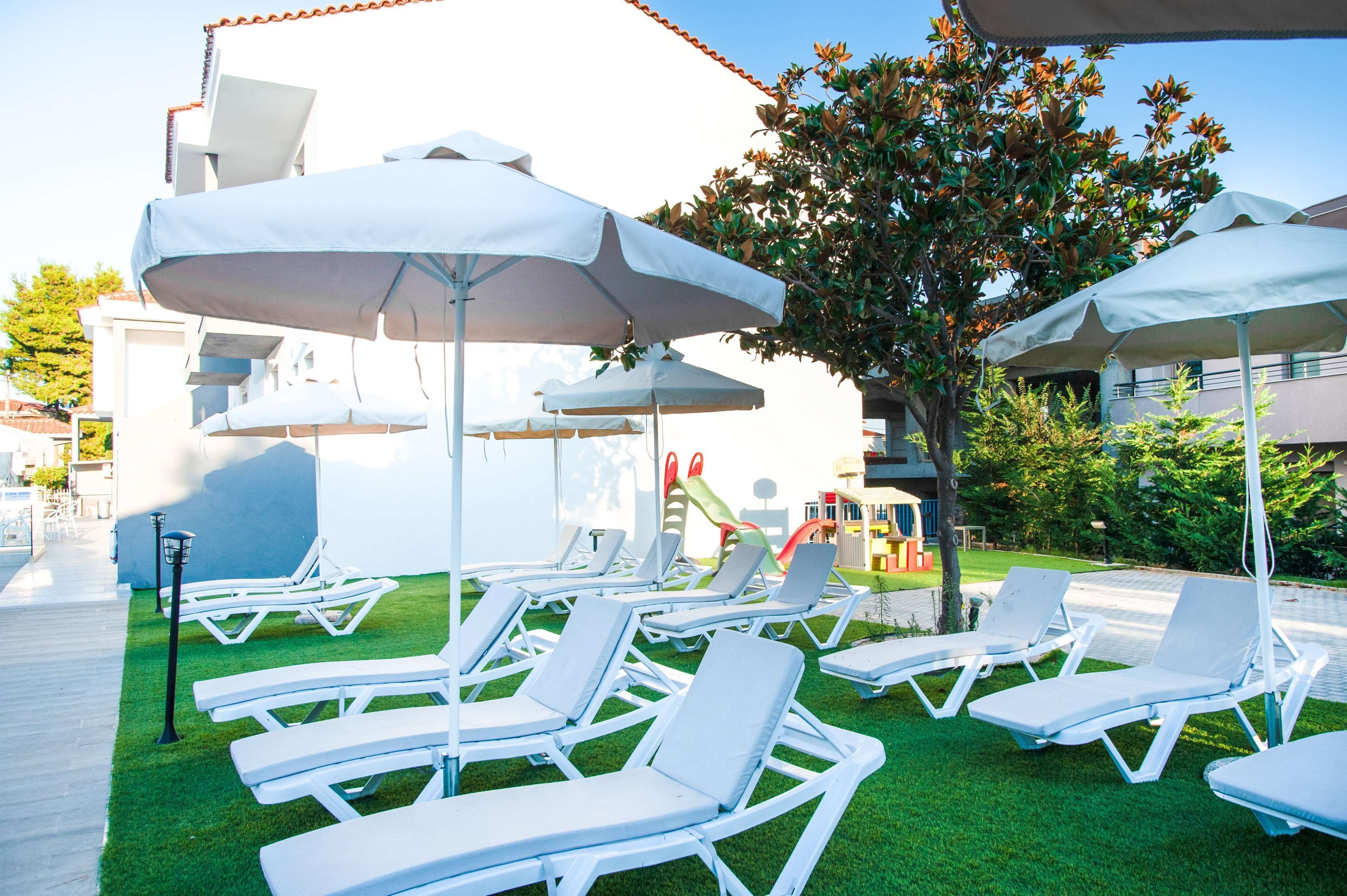 Uma área de piscina com espreguiçadeiras e guarda-sóis brancos, um espaço de recreação infantil e um edifício com varandas.