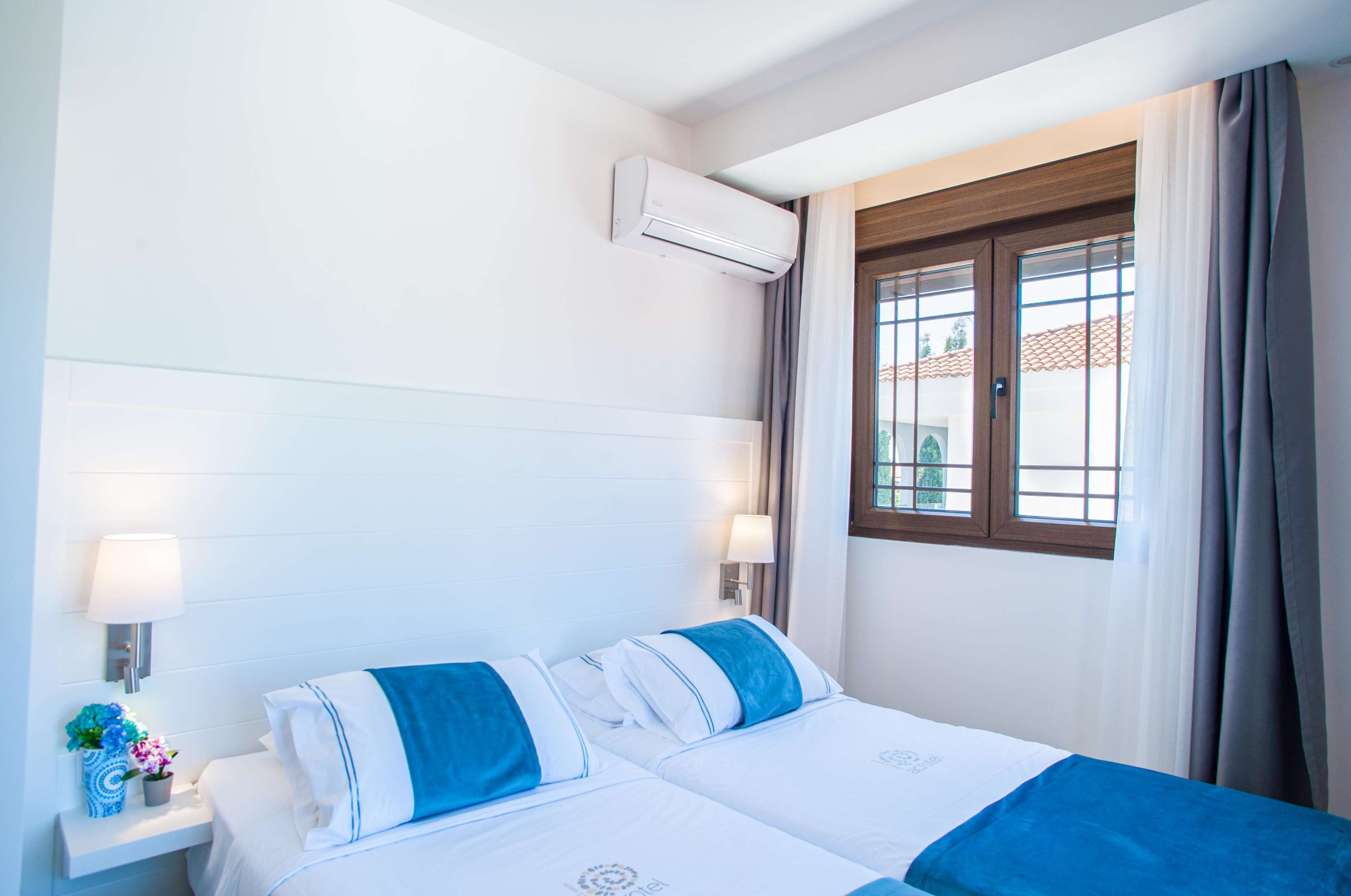 Quarto de hotel com duas camas, uma janela com cortinas, ar condicionado e uma mesa de cabeceira com abajur e flores.