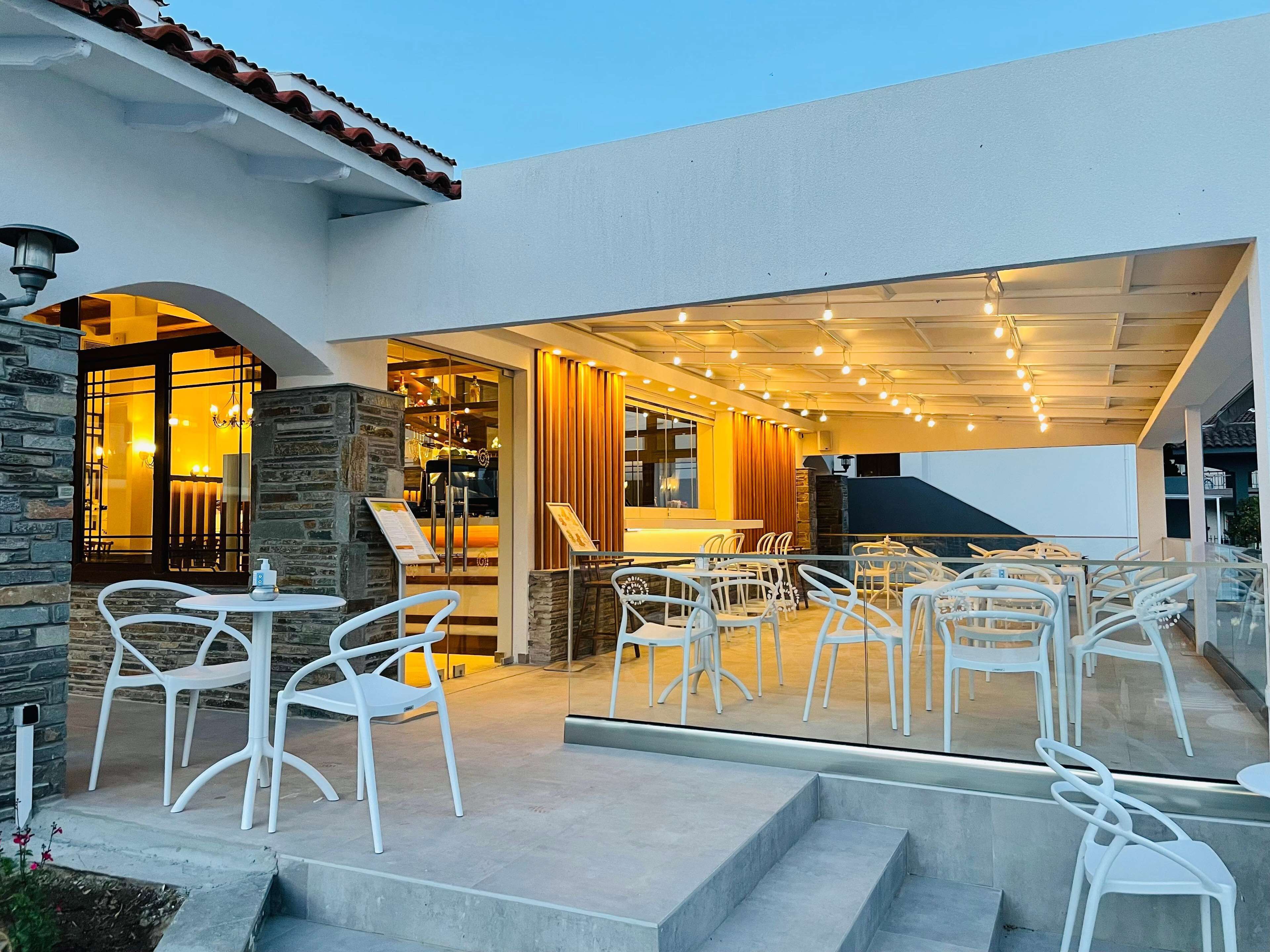 Um pátio externo com cadeiras e mesas brancas, uma parede de pedra e o interior de um restaurante visível através de uma porta de vidro.