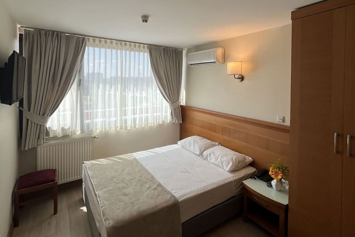 Quarto de hotel com cama, mesa de cabeceira, guarda-roupa, radiador e janela com cortinas.