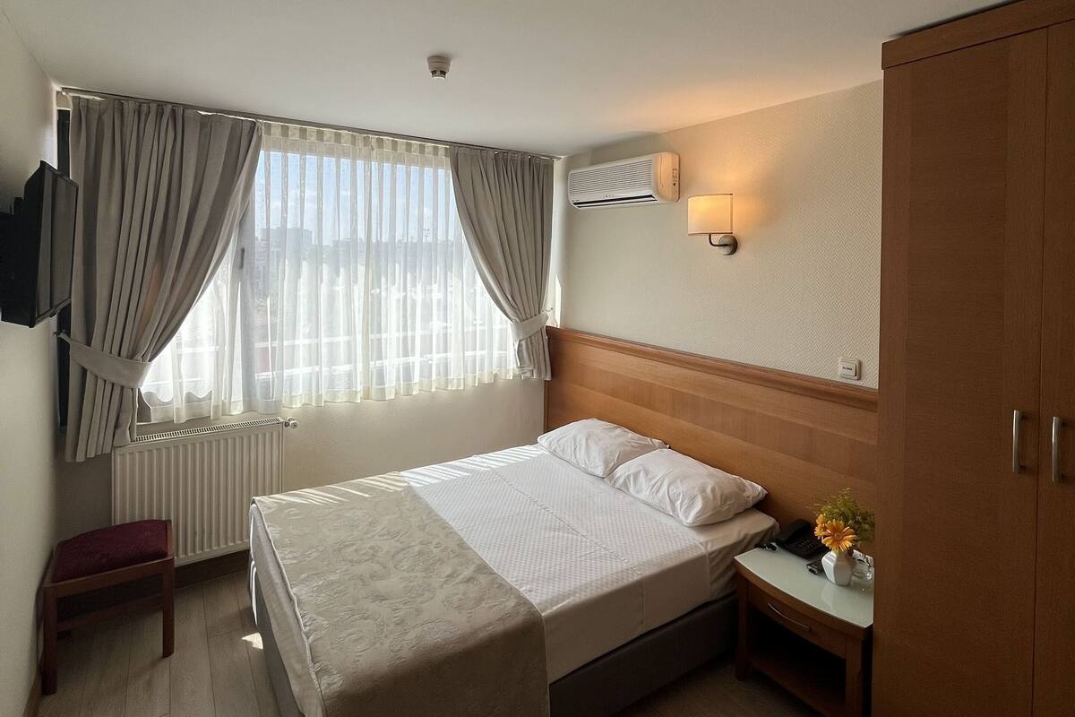 Quarto de hotel com cama, mesa de cabeceira com luminária, armário, radiador e janela com cortinas.