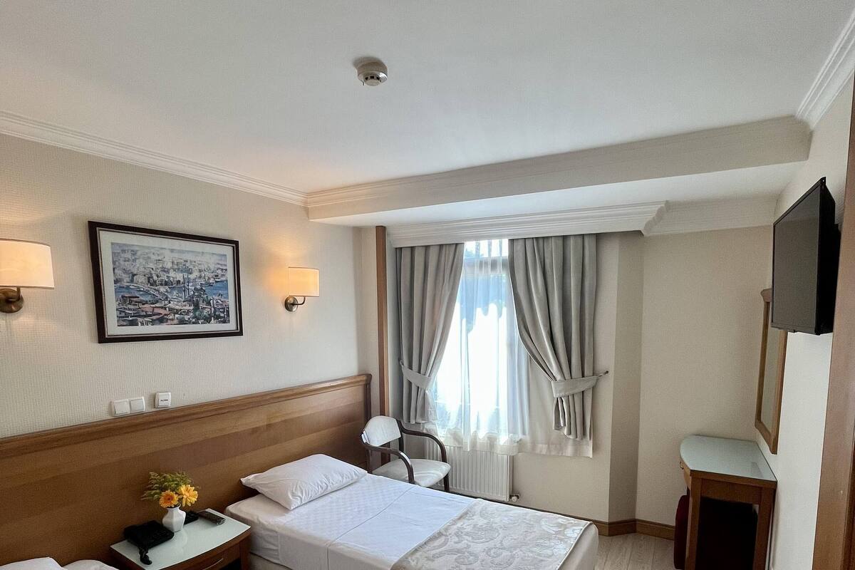 Quarto de hotel com duas camas, uma escrivaninha com cadeira, televisão e janela com cortinas.