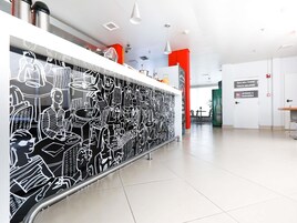 Um hall de hotel moderno com um balcão de recepção alongado, um pilar vermelho e uma placa indicando acesso à garagem.