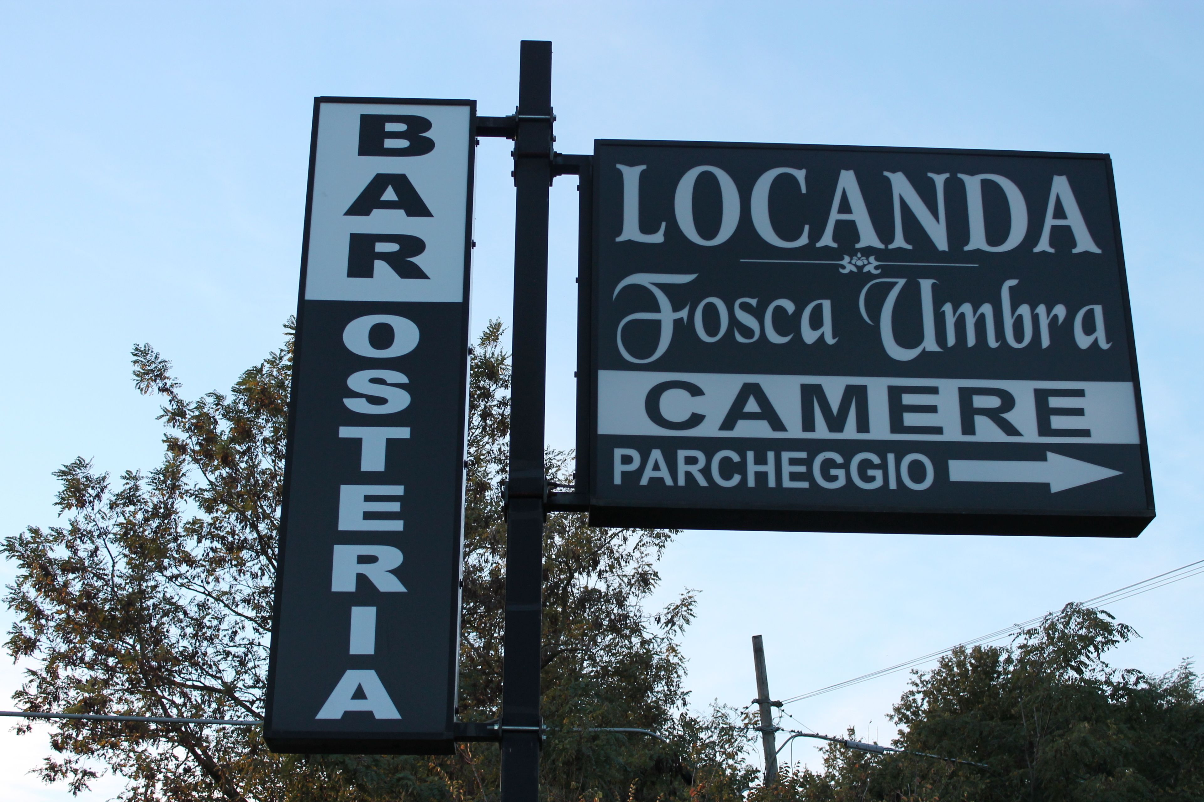 Uma placa de direção com 'BAR OSTERIA' e 'LOCANDA FoscA Umbra CAMERE PARCHEGGIO' apontando para a direita.