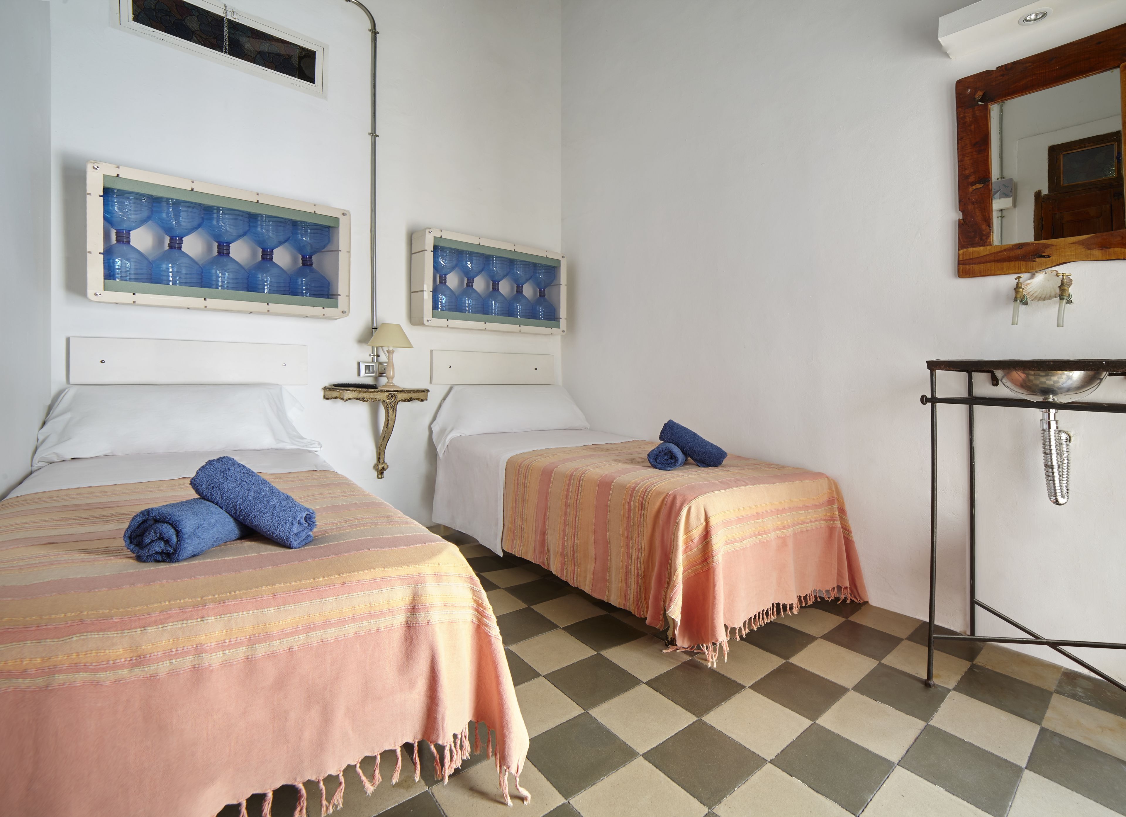 Quarto de hotel com duas camas, piso xadrez, pia com espelho e bebedouros fixados na parede.