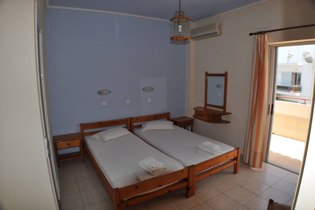 Uma cama de casal com duas almofadas e uma mesa de cabeceira, um penteou de madeira, um espelho e uma porta de correr para uma varanda.