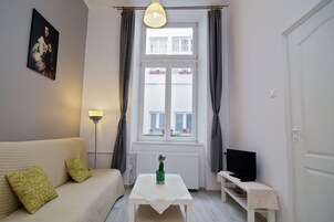 Quarto com sofá branco, uma mesinha com planta, televisão em móvel e janela com cortinas.