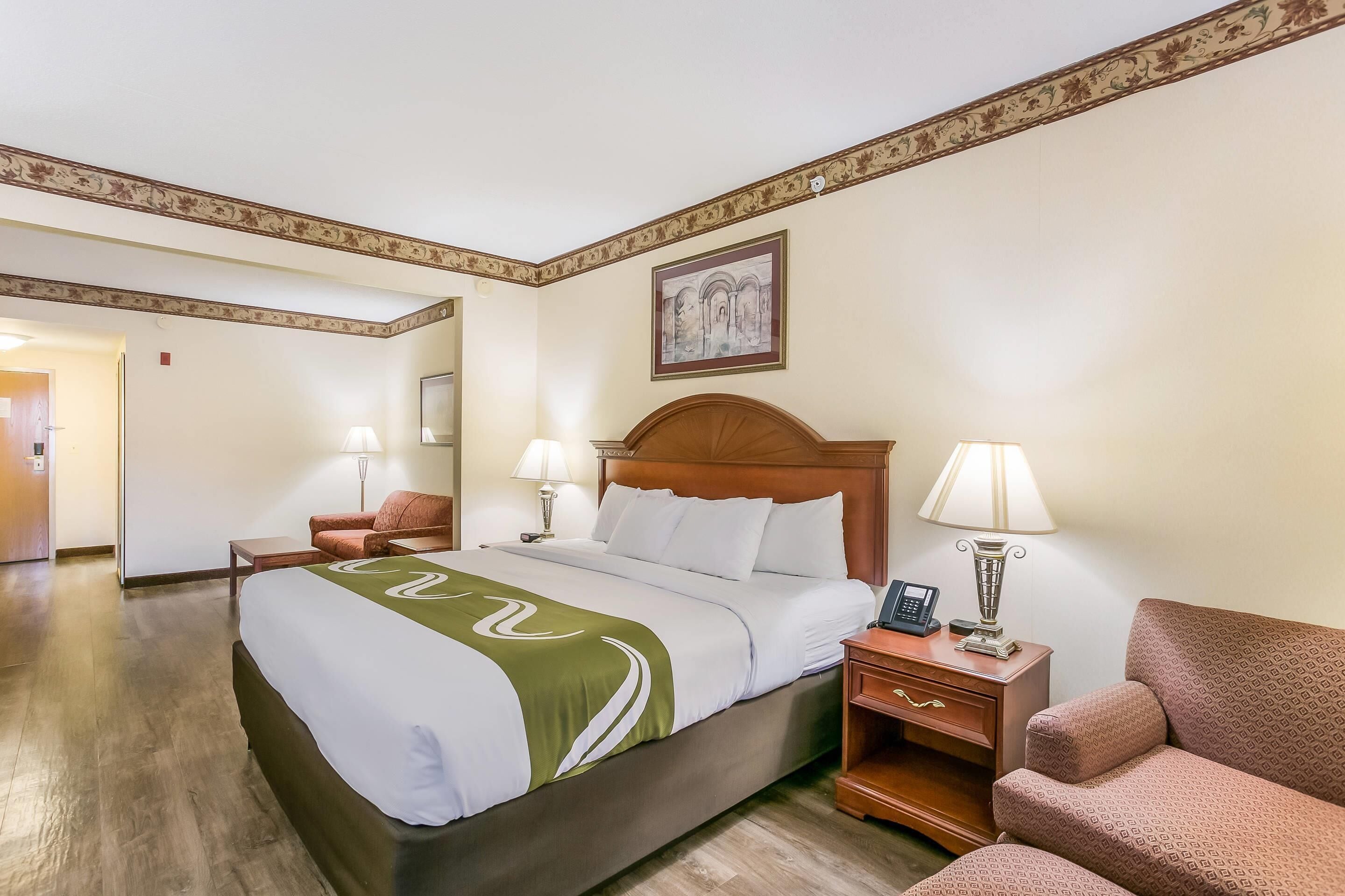 Quarto de hotel com uma cama grande, duas luminárias de cabeceira, uma mesinha de cabeceira de madeira, uma poltrona marrom e uma obra de arte emoldurada na parede.