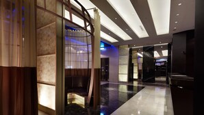 Um hall de hotel moderno com piso de mármore, área de estar com divisória de vidro e teto com iluminação embutida.