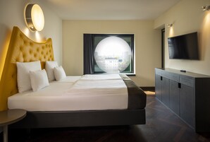 Quarto de hotel moderno com uma cama grande, televisão de tela plana, espelho redondo e vista para o horizonte urbano através de uma janela ampla.
