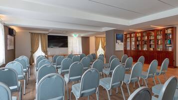 Uma sala de conferências com fileiras de cadeiras, uma tela grande e uma estante de livros.