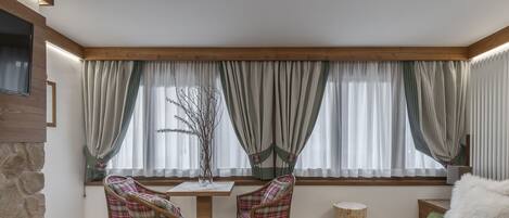 Um quarto acolhedor com um banco de madeira, duas cadeiras xadrez, uma mesinha e uma janela grande com cortinas leves.