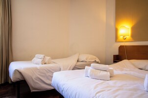 Duas camas de solteiro com roupa de cama e toalhas brancas, uma cabeceira de madeira e um luminário fixado na parede.