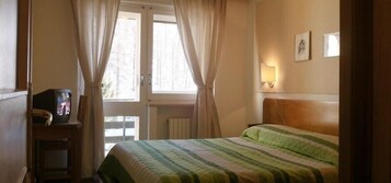 Quarto de hotel com cama, escrivaninha com luminária e janela com cortinas.