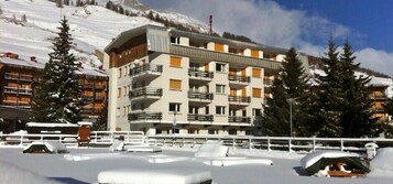 Um hotel com várias andares e varandas, cercado por montanhas e árvores cobertas de neve.