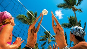 Jogo de voleibol de praia com jogadores esticando-se para a bola, palmeiras e céu azul.