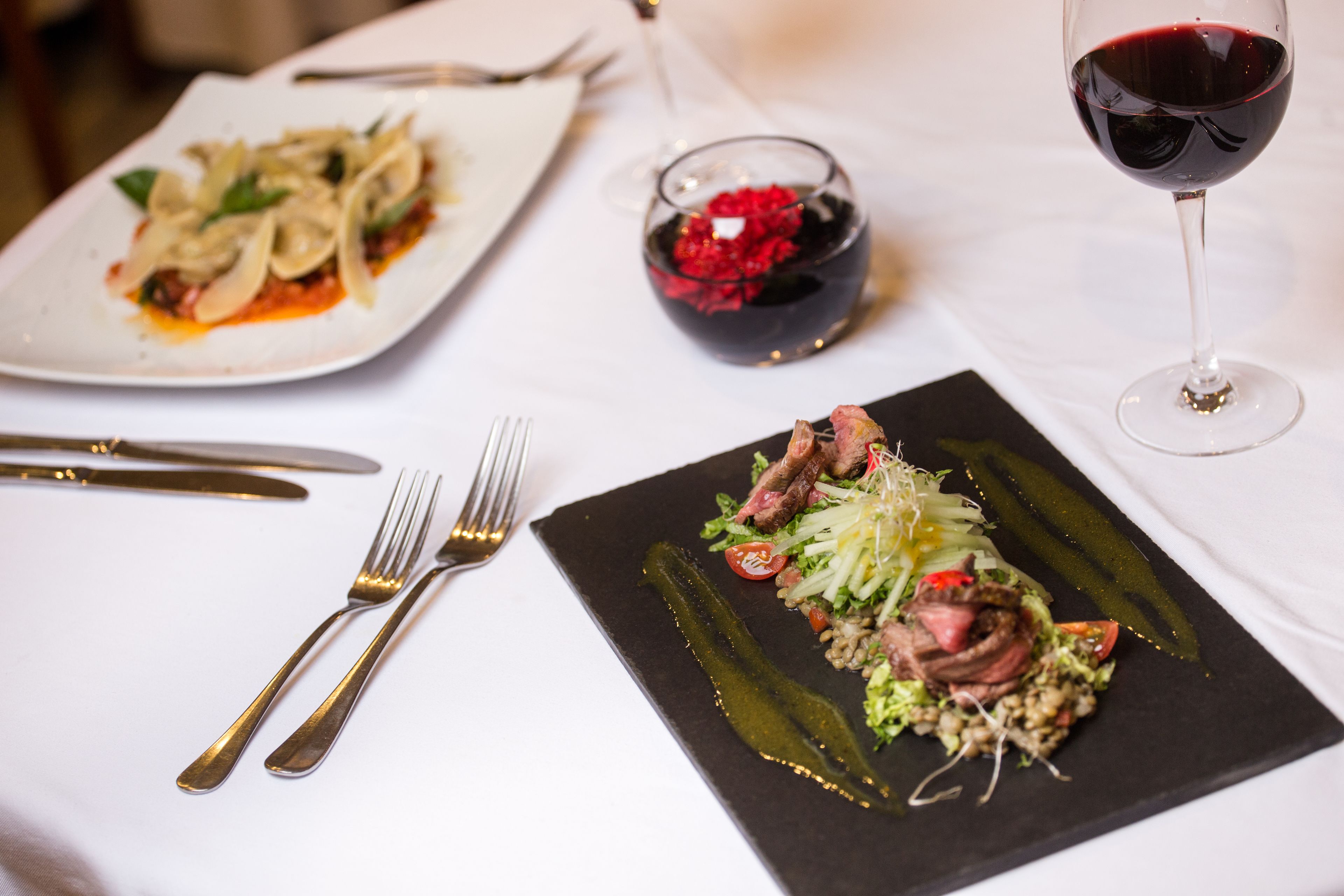 Uma mesa de jantar com um prato de massa, um copo de vinho tinto e um prato preto com um prato de carne.