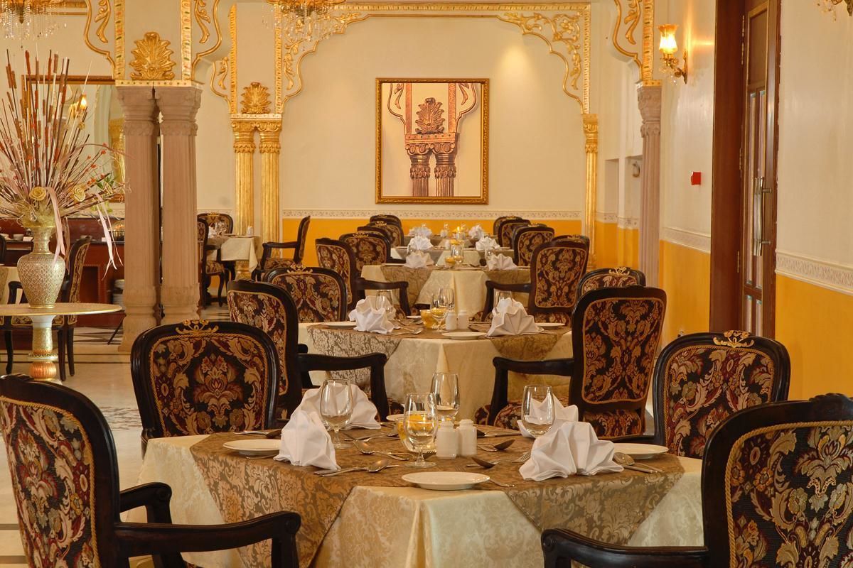 Uma sala de jantar formal com cadeiras ornamentadas, mesas postas para refeição e um candelabro.