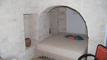 Um quarto pequeno e rústico com parede de pedra, uma cama simples com colcha estampada, uma mesinha com um livro e uma cadeira com almofada vermelha.