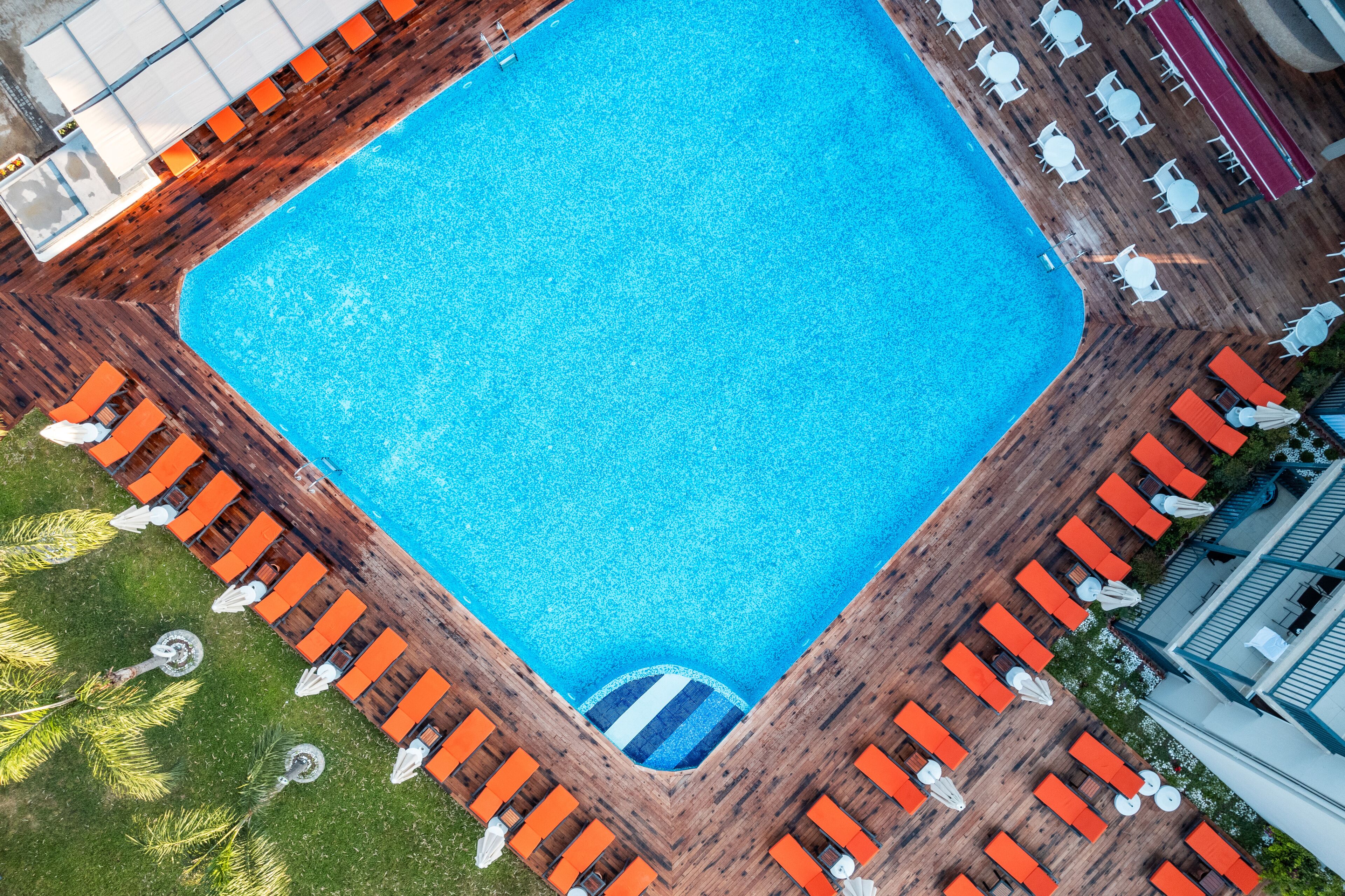 Vista aérea de uma piscina retangular rodeada por cadeiras de banho laranja e guarda-sóis brancos, com um deck de madeira e uma pequena área circular com padrão azul e branco listrado.