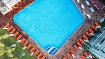 Vista aérea de uma piscina retangular rodeada por cadeiras de banho laranja e guarda-sóis brancos, com um deck de madeira e uma pequena área circular com padrão azul e branco listrado.