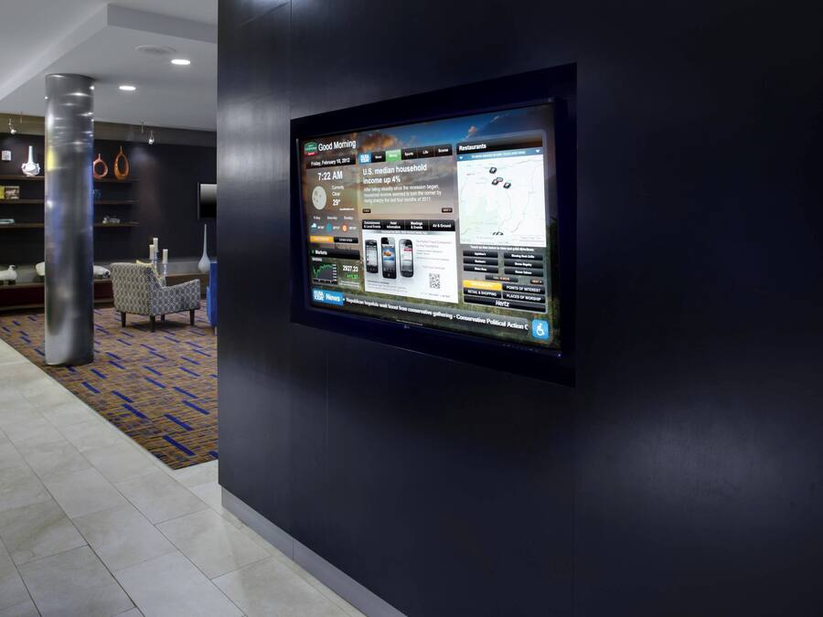 Um hall moderno com um painel digital exibindo notícias e previsão do tempo, um balcão de recepção e um carpete estampado.