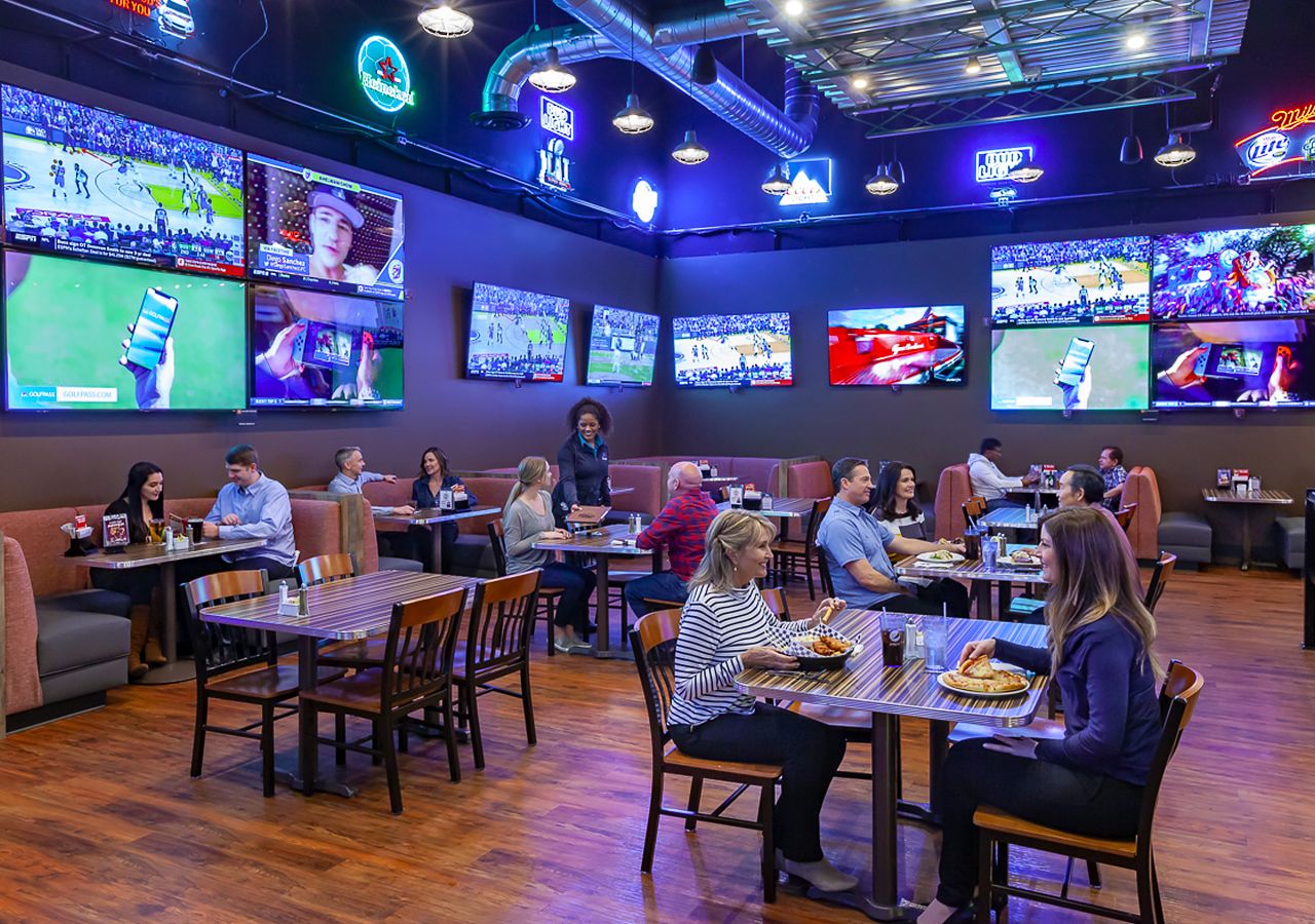 Um restaurante com várias telas de TV exibindo eventos esportivos, mesas e cadeiras de madeira, e um ambiente descontraído para refeições.