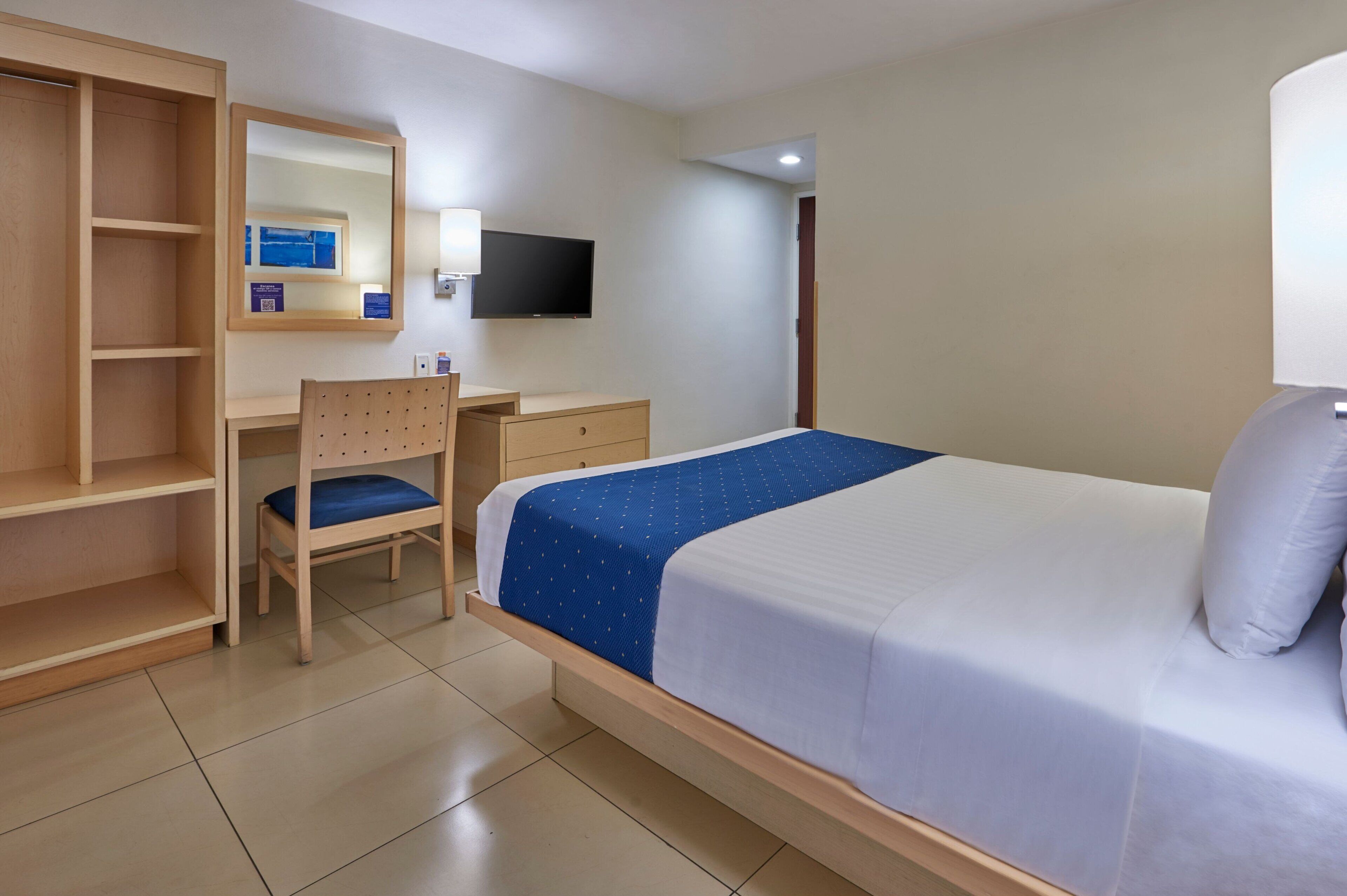 Quarto de hotel com cama, escrivaninha, cadeira, guarda-roupa, televisão e espelho.