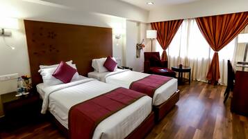 Quarto de hotel com duas camas, piso de madeira, uma escrivaninha, uma cadeira e uma janela com cortinas.