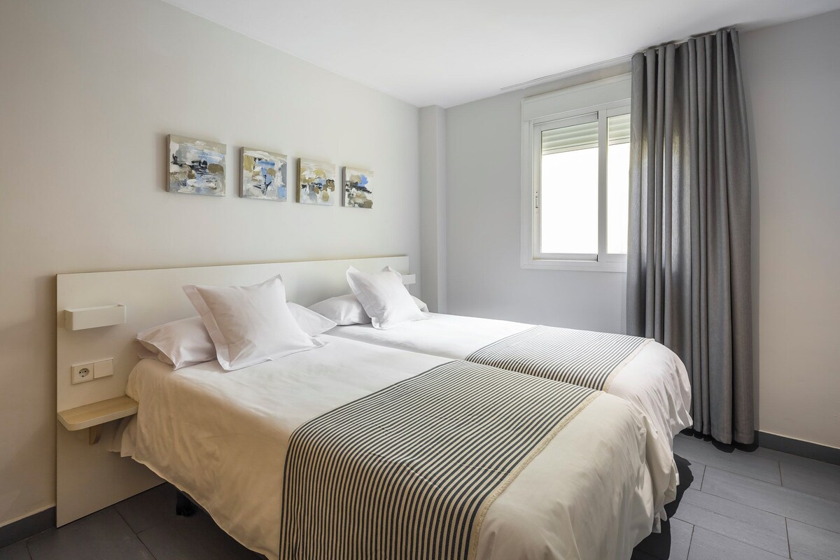 Cama de casal com roupa de cama branca e corredores listrados, mesa de cabeceira e criado-mudo. Há três obras de arte penduradas na parede acima da cama.
