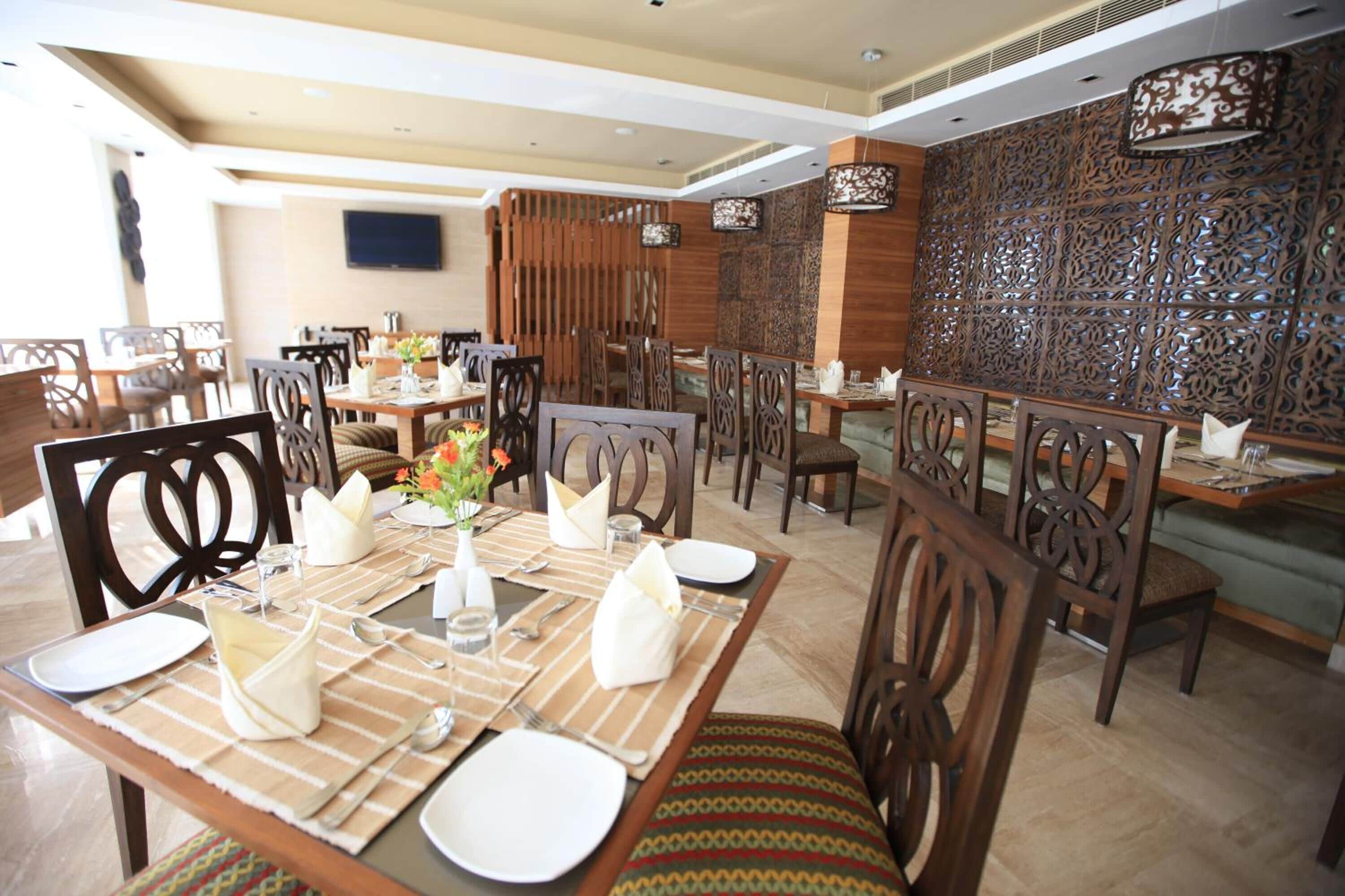 Uma sala de jantar com várias mesas preparadas para o serviço, cadeiras de madeira e um painel decorativo na parede.