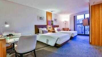 Quarto de hotel com duas camas, uma escrivaninha com cadeira, televisão e uma janela grande com vista para a cidade.