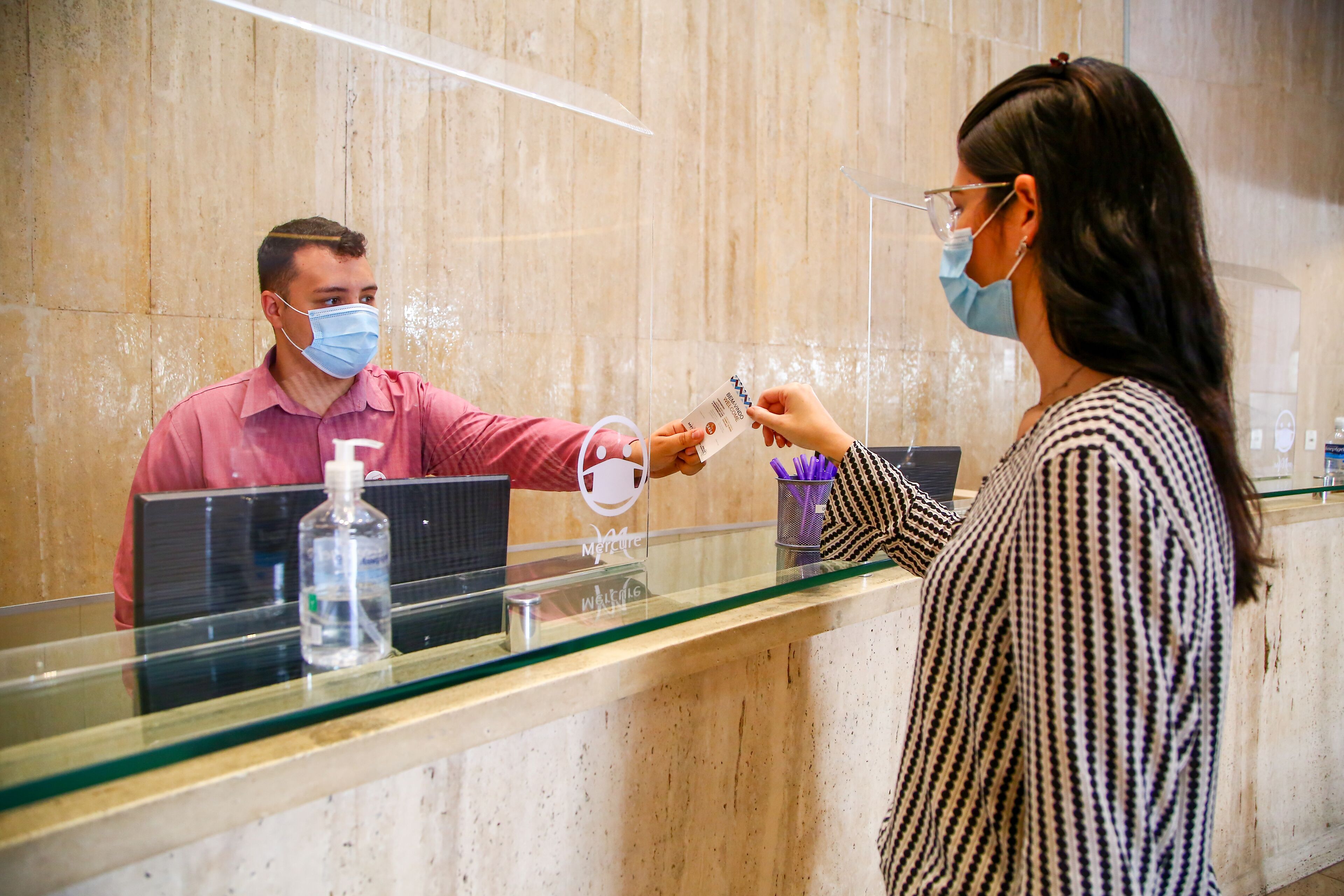 Uma receção com divisória de vidro, um frasco de álcool em gel e uma máscara facial no balcão.