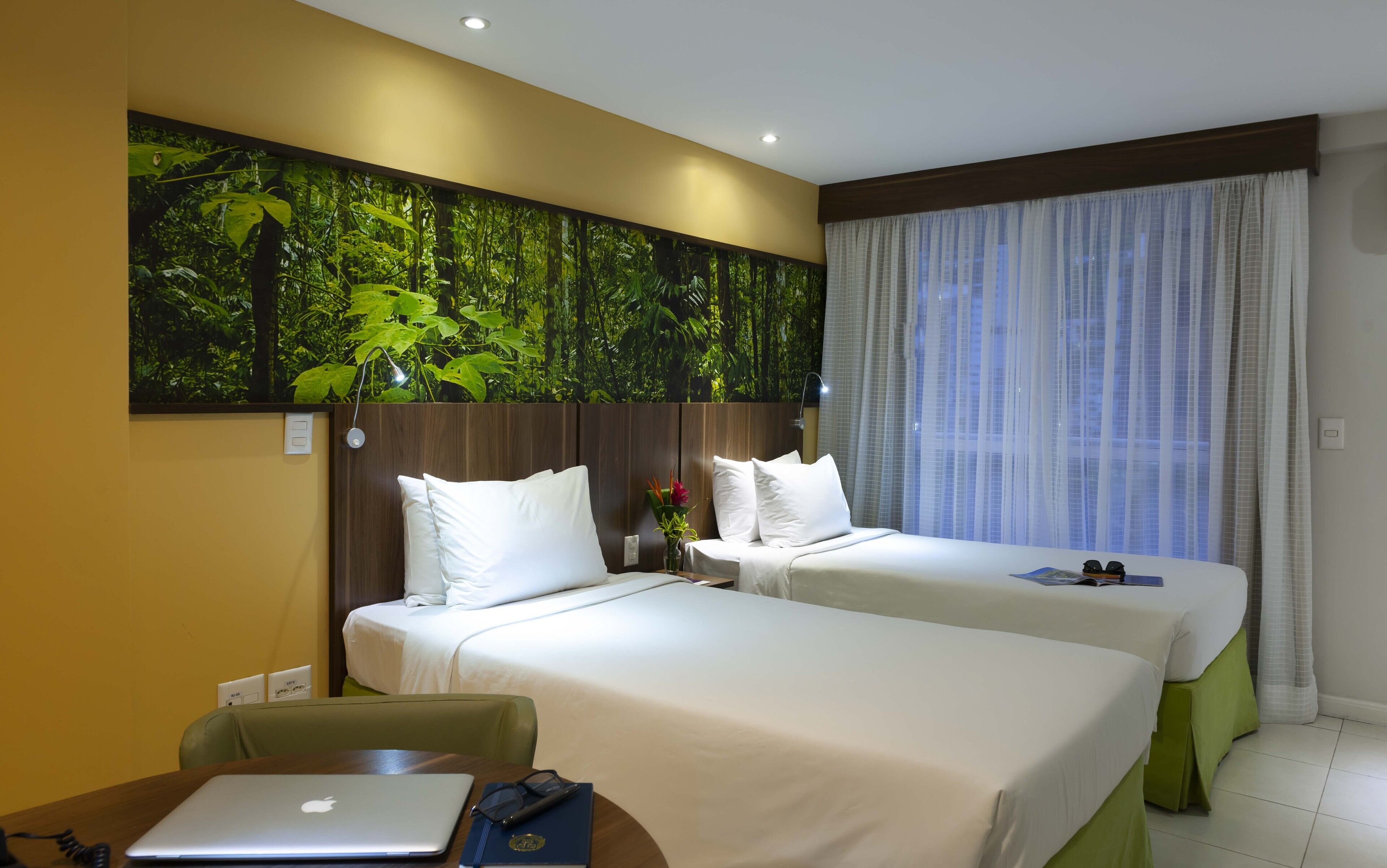 Quarto de hotel com duas camas, uma escrivaninha com um laptop, uma cadeira e um mural grande na parede retratando uma floresta.