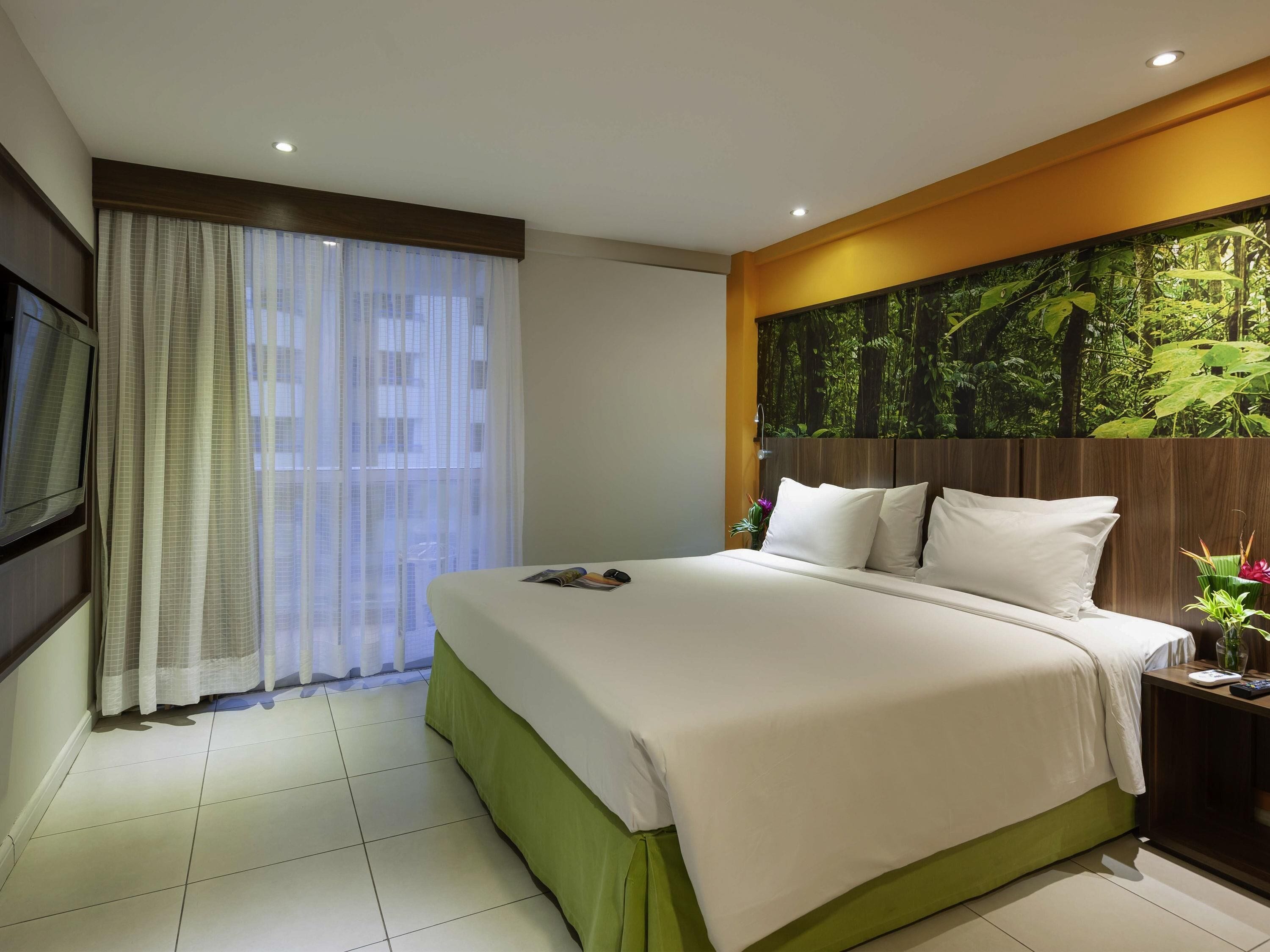 Quarto de hotel com cama, TV fixada na parede, janela com cortinas translúcidas e um mural na parede com tema de floresta.