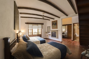 Quarto com duas camas, vigas de madeira no teto, lareira e um espaço de refeições ao fundo.