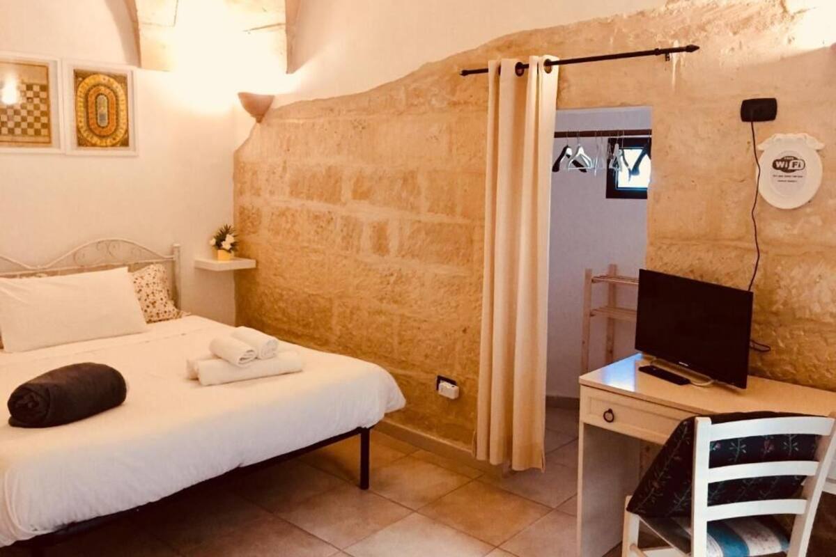 Um quarto com uma cama, uma escrivaninha com um computador, uma TV e uma cortina.