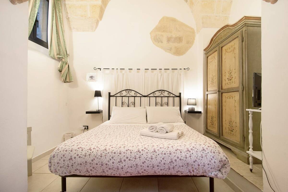 Quarto com cama, duas mesas de cabeceira com abajures, guarda-roupa e parede de pedra.