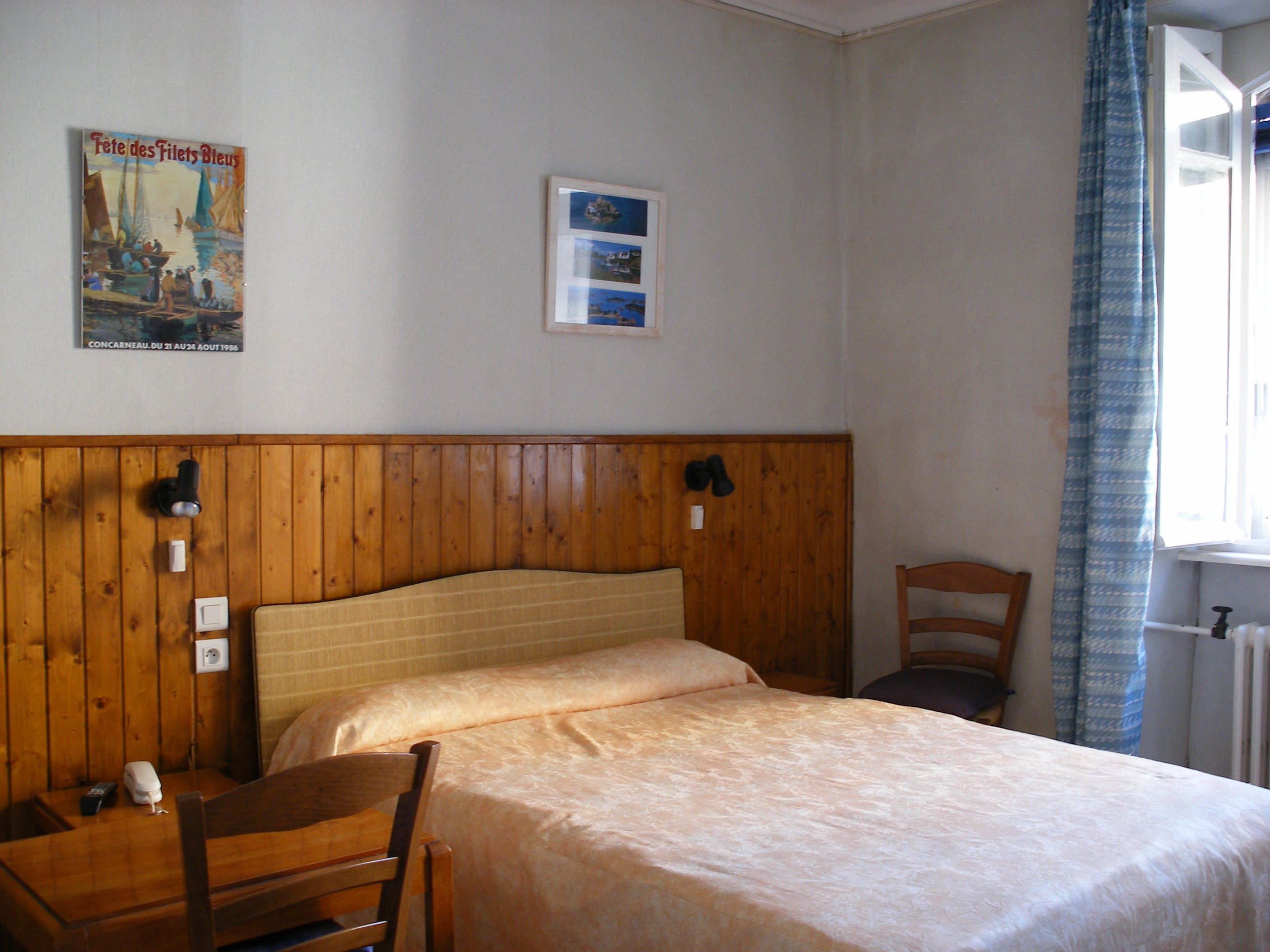 Quarto com cabeceira de madeira, cama com colcha bege, cadeira de madeira, mesa pequena com controle remoto e um pôster na parede.