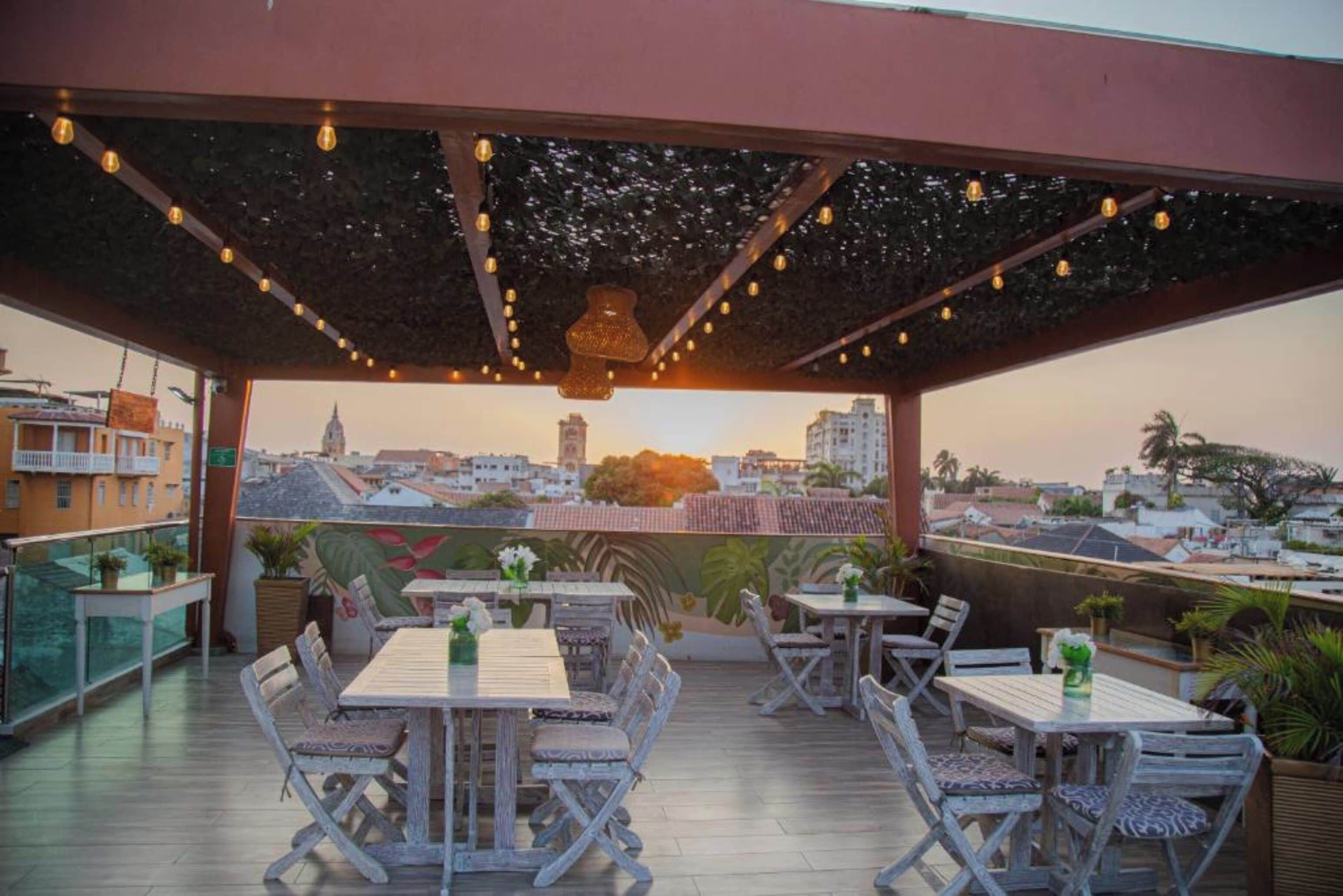 Um pátio externo com mesas e cadeiras, um mural na parede e vista para o horizonte da cidade.