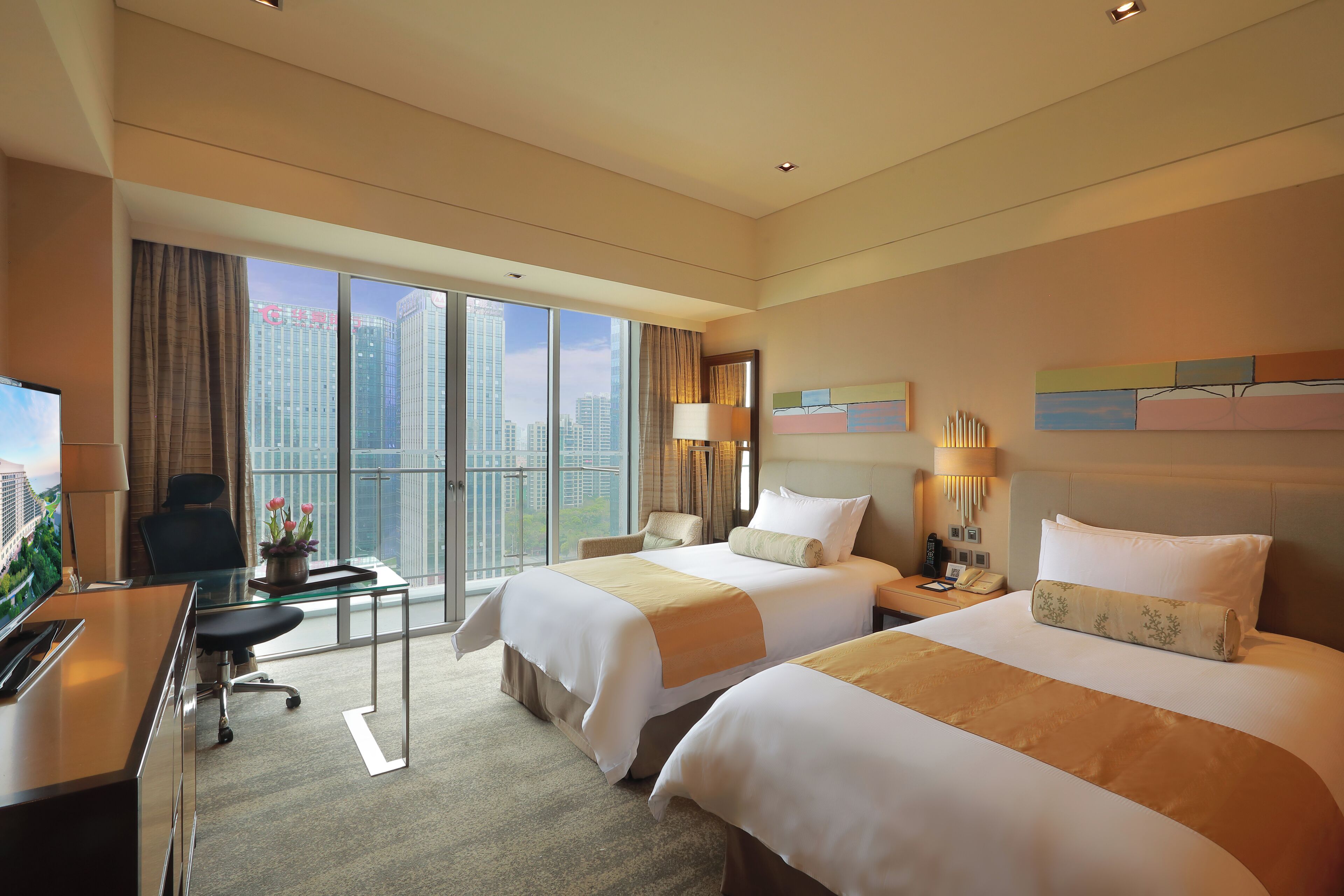 Quarto de hotel com duas camas, uma escrivaninha, uma cadeira, uma televisão e vista para a paisagem urbana.