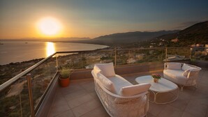 Uma varanda com móveis de jardim brancos, um vaso com planta e vista para o mar ao pôr do sol.