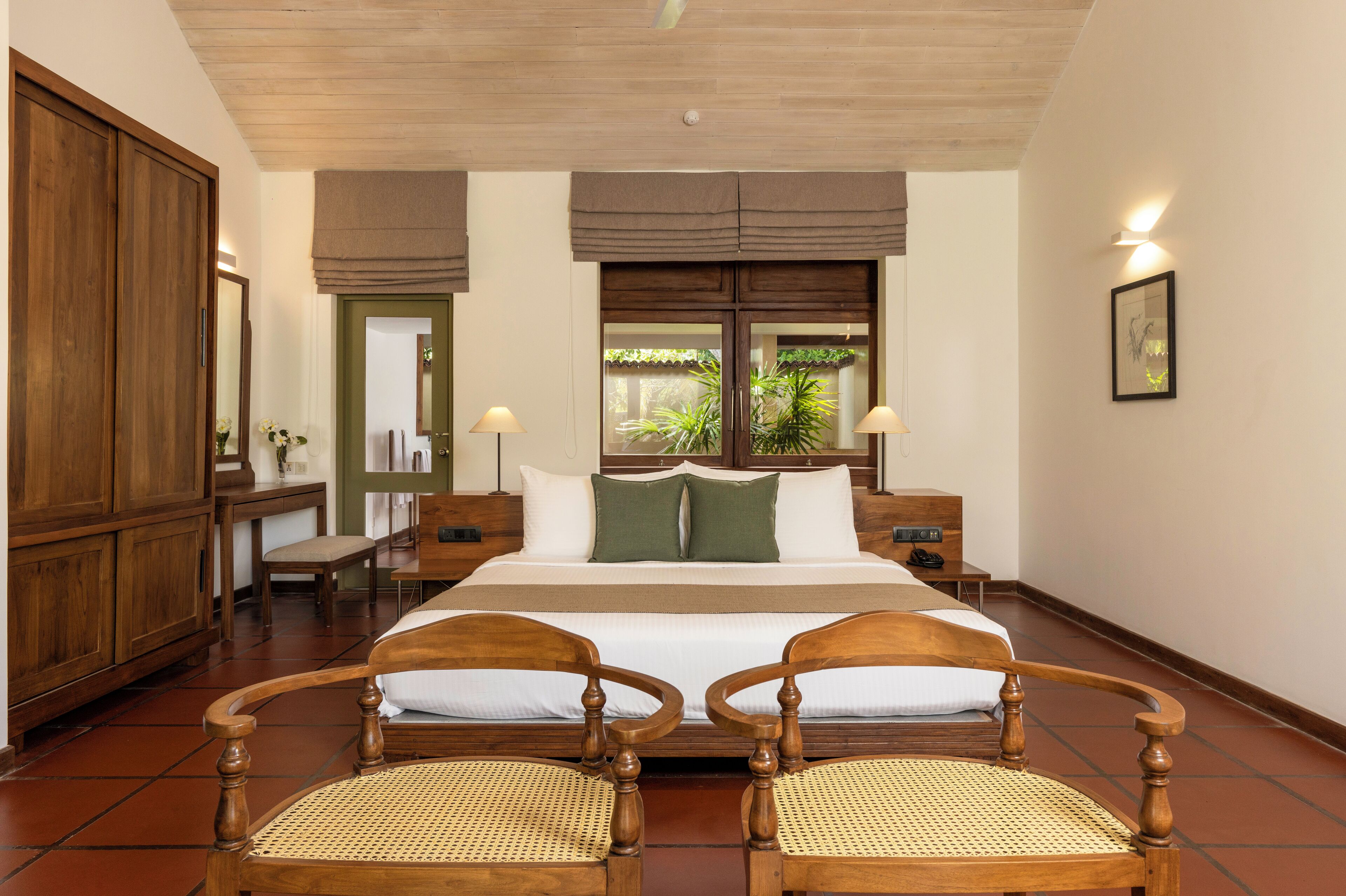 Quarto de hotel com uma cama grande, duas cadeiras de madeira, uma mesa de cabeceira e uma janela com vista para o verde.