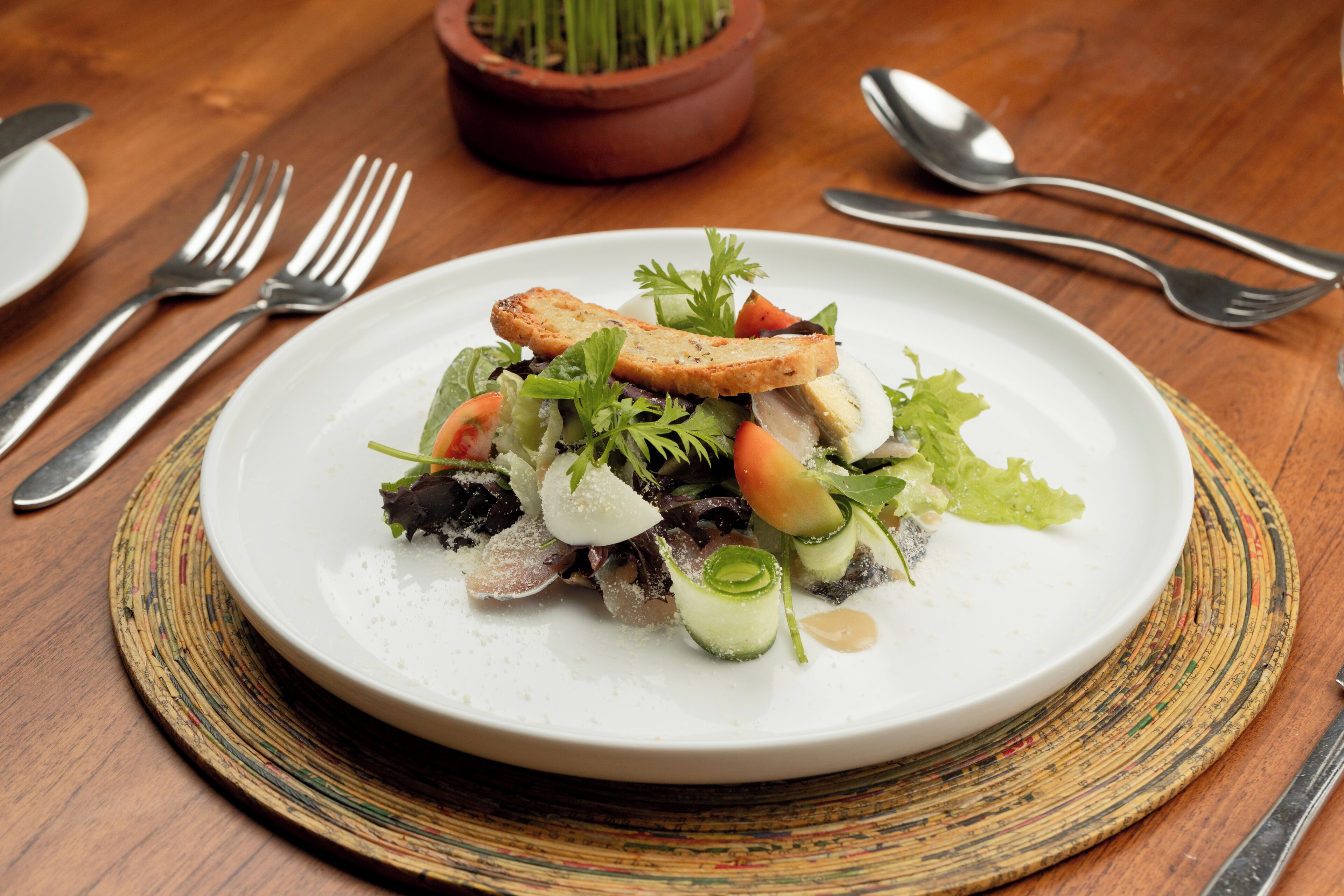 Uma salada com vários vegetais e uma fatia de pão em um prato branco, sobre uma mesa de madeira com um sousplat trançado.