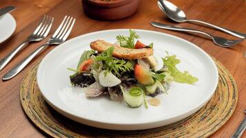 Uma salada com vários vegetais e uma fatia de pão em um prato branco, sobre uma mesa de madeira com um sousplat trançado.