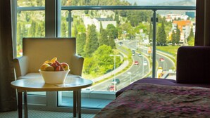 Quarto de hotel com uma janela ampla que oferece vista para uma estrada e vegetação. Há uma cadeira com uma tigela de frutas sobre uma mesinha.