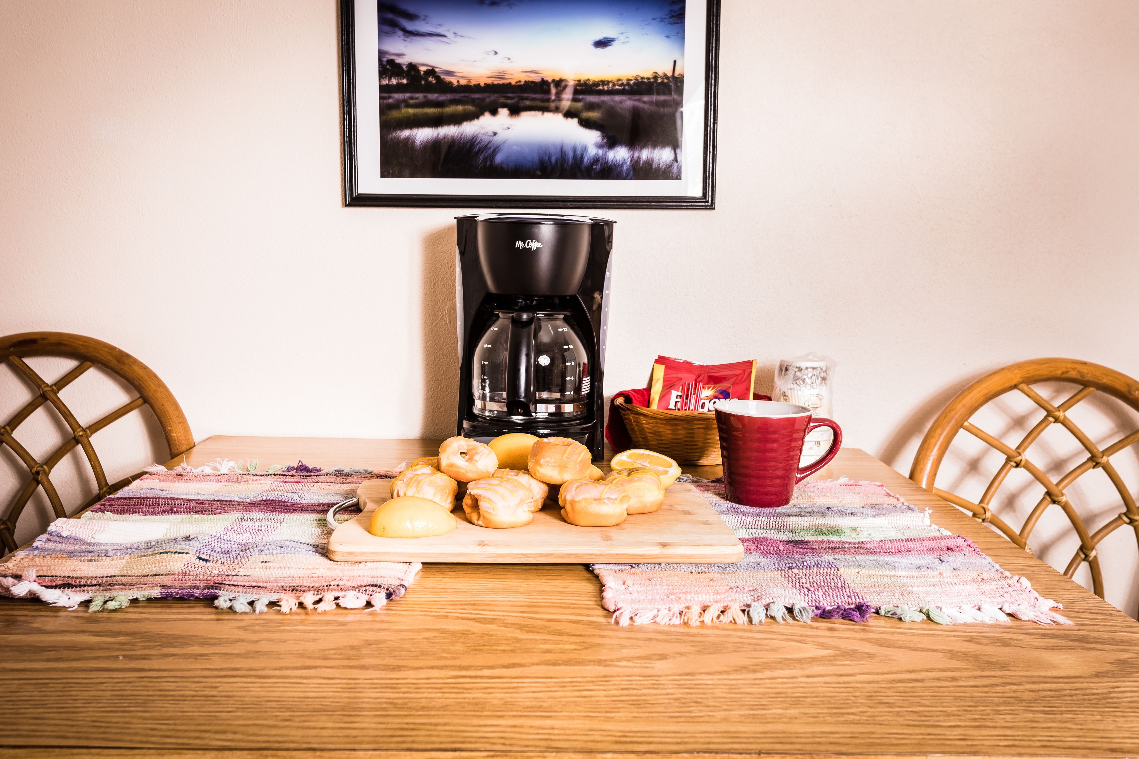 Uma mesa de madeira com uma cafeteira, uma cesta de pão e uma caneca vermelha.